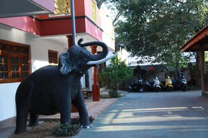 Uma estátua de elefante em frente a um edifício com fachada vermelha e branca, um caminho de azulejos e scooters estacionadas ao fundo.