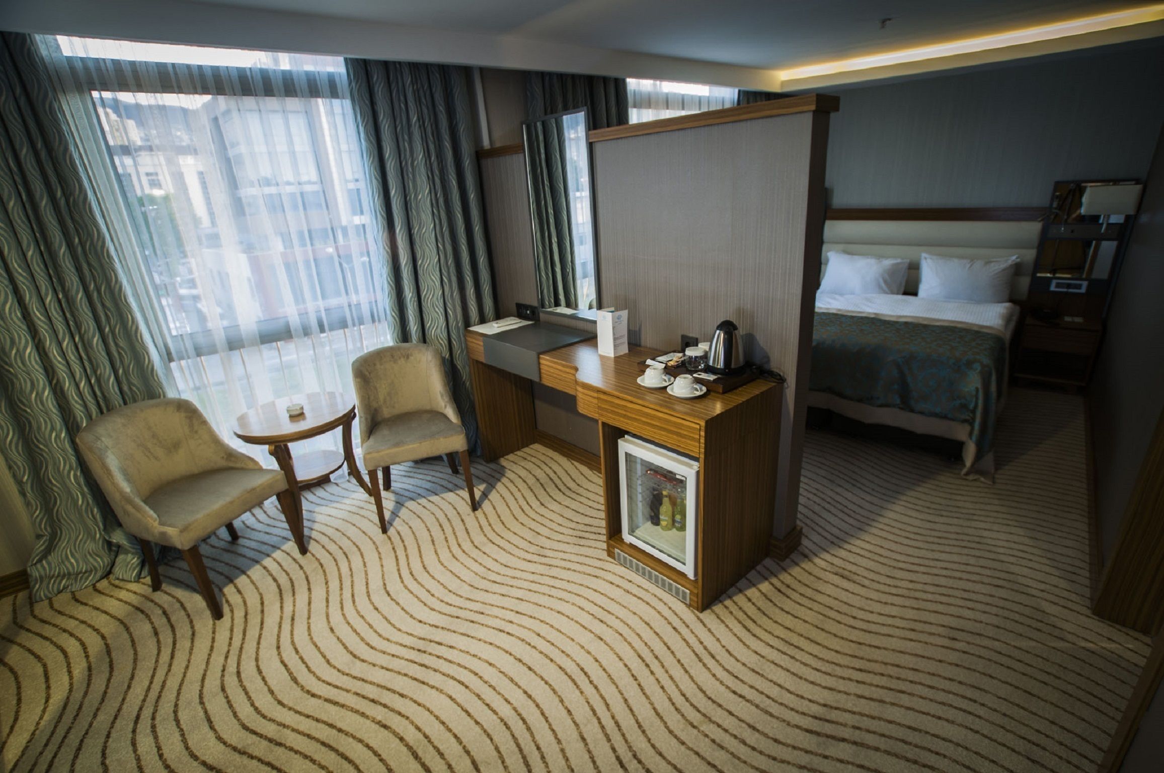 Quarto de hotel com cama, secretária com computador, duas cadeiras, minibar e vista da cidade por grandes janelas.