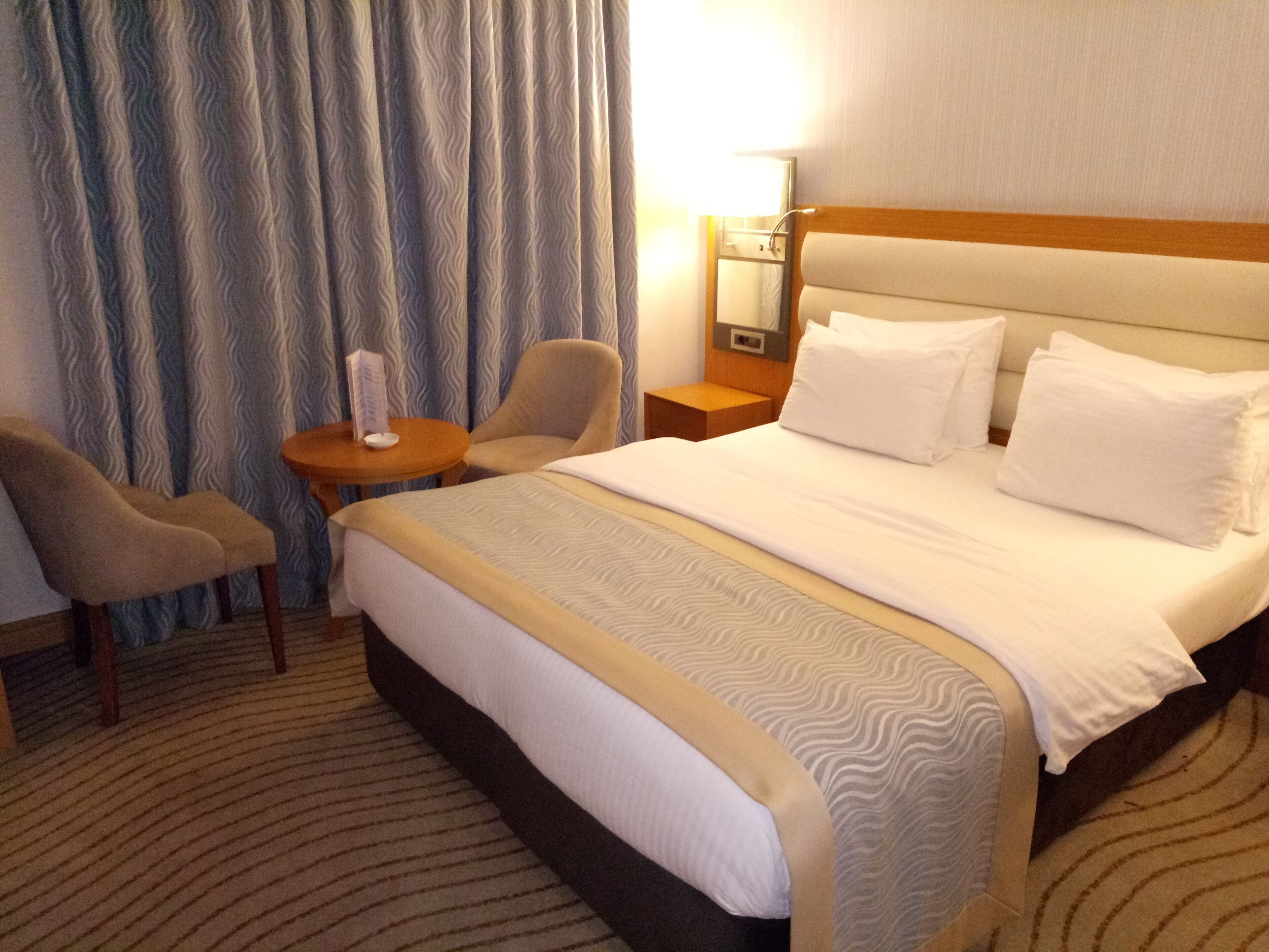 Quarto de hotel com uma cama grande, uma mesinha de cabeceira, uma cadeira e um abajur.