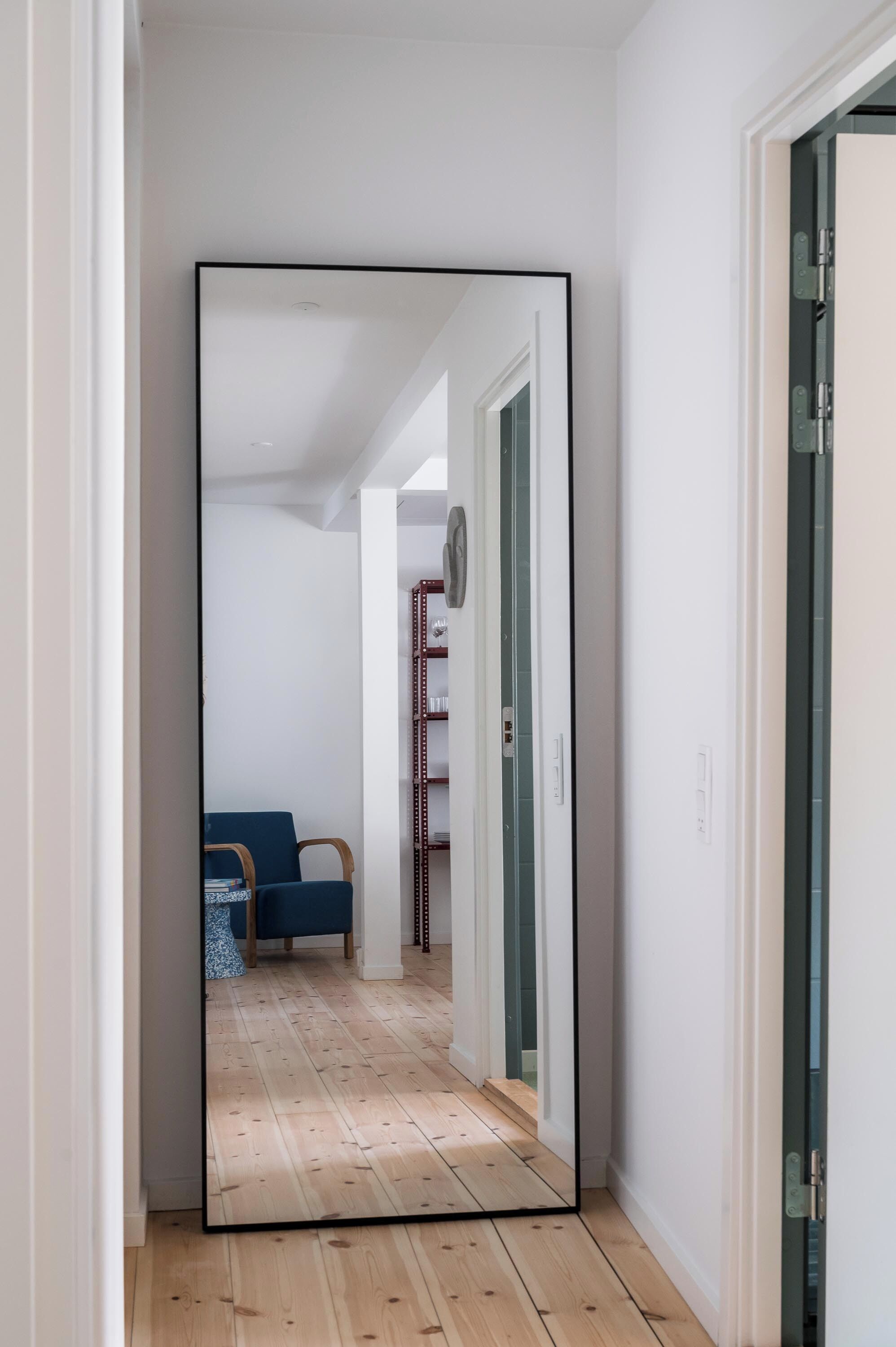 Um corredor estreito com um espelho grande refletindo um quarto com uma cadeira azul e uma estante de livros.