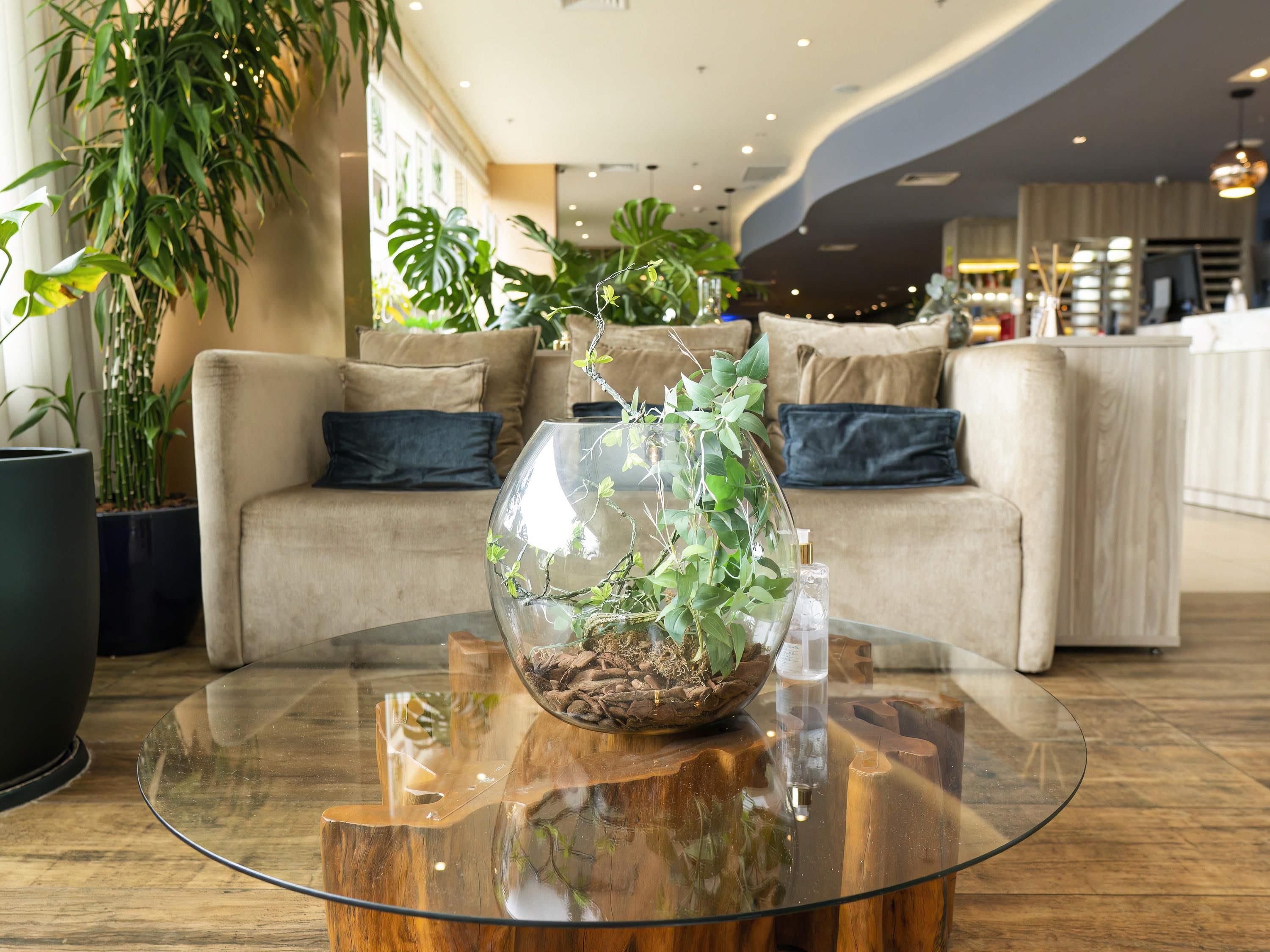 Uma sala de estar moderna com uma mesa de centro de vidro, um sofá bege e diversas plantas de interior.