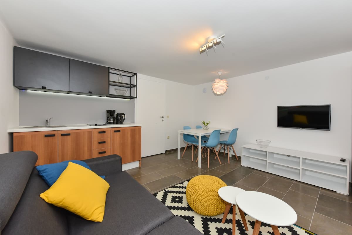 Uma sala de estar moderna com um sofá cinza, almofadas amarelas e azuis, um tapete com padrão preto e branco, uma mesinha branca e um banquinho amarelo.