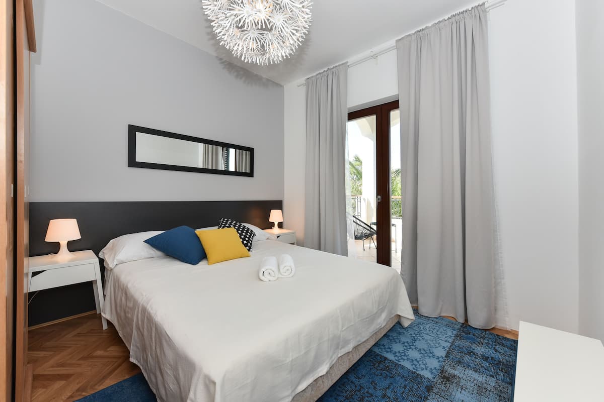 Quarto de hotel com uma cama grande, uma mesa de cabeceira com abajur, luminária no teto e vista para o exterior através de uma porta de correr de vidro.