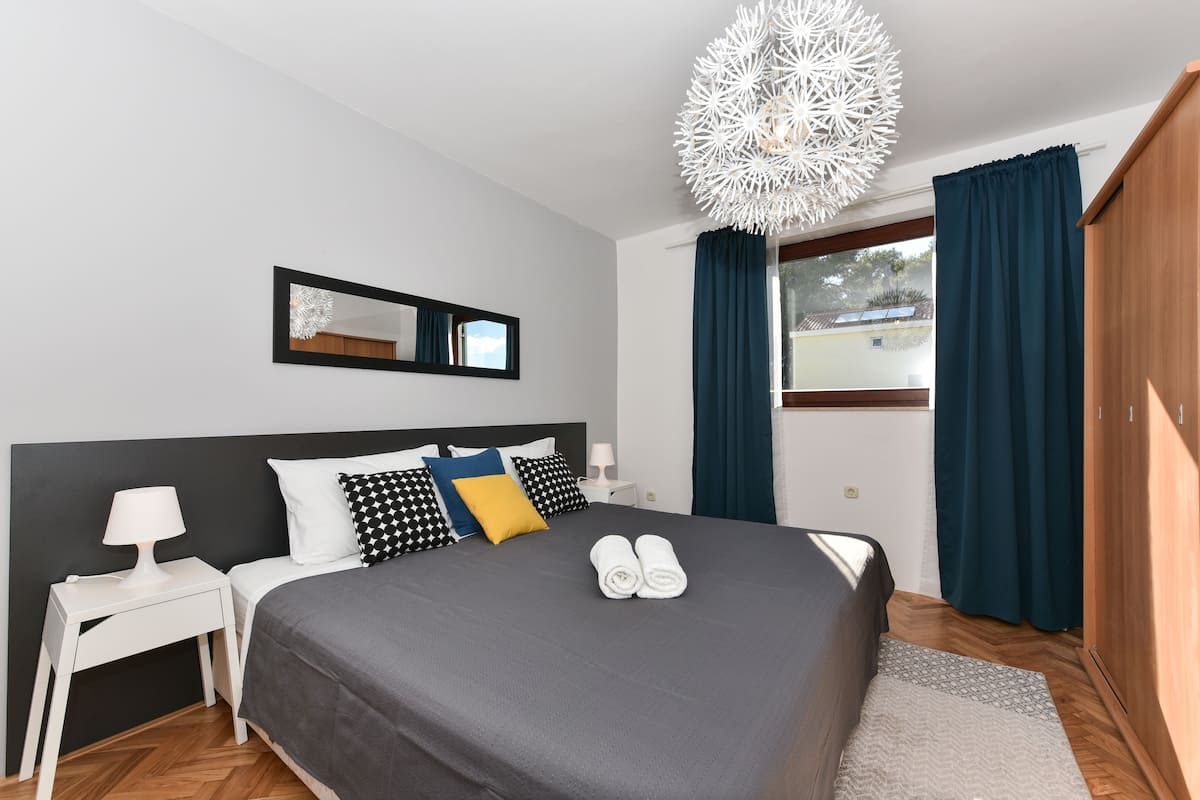 Um quarto moderno com uma cama grande, uma mesa de cabeceira com abajur, um espelho e vista para o exterior através de uma janela com cortinas.