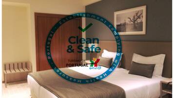 Quarto de hotel com cama, mesa de cabeceira e cadeira. Há um logótipo com o texto