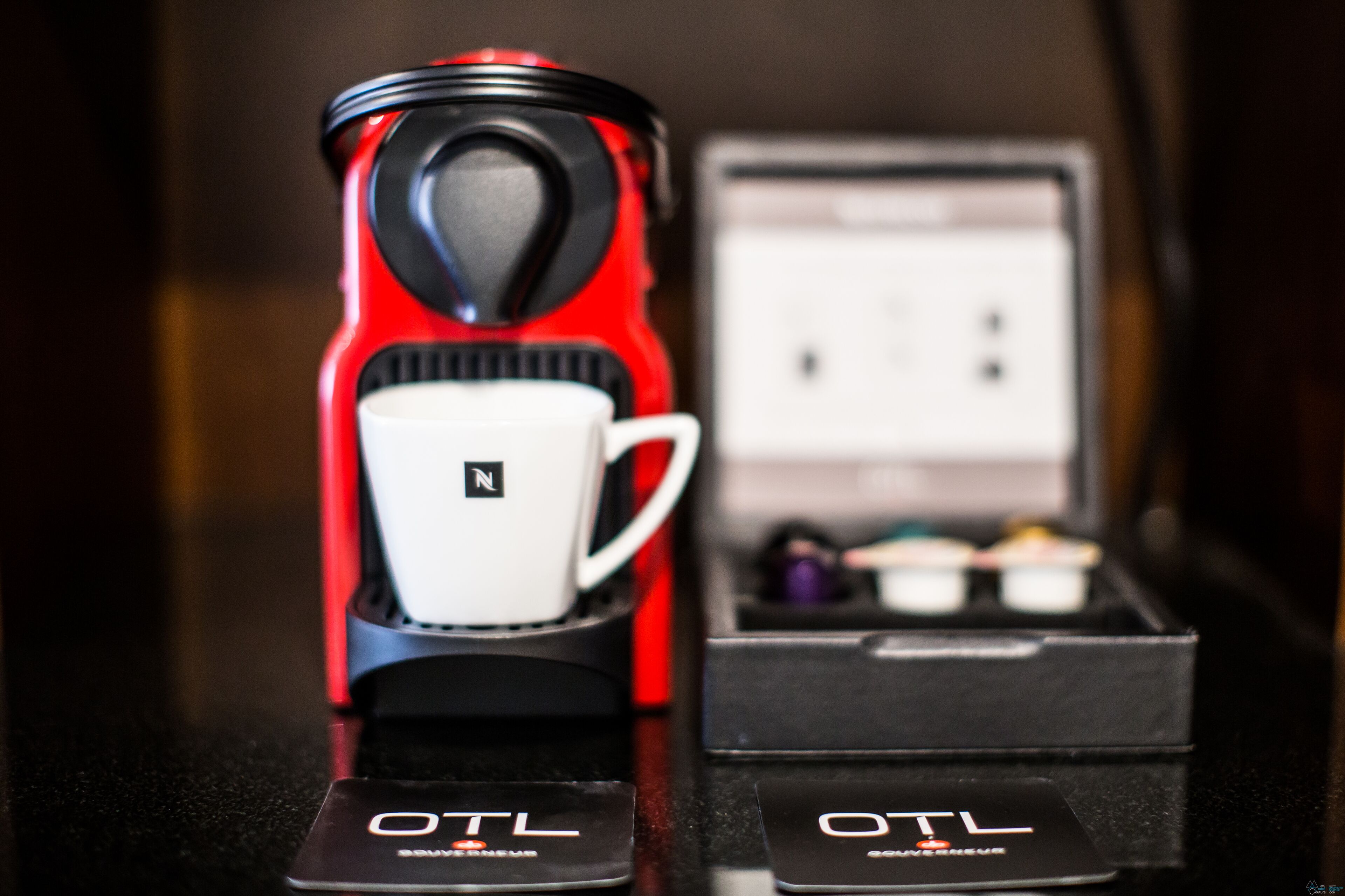 Uma máquina de café com uma xícara branca, duas caixas pretas com a etiqueta "OTL" e um fundo desfocado.