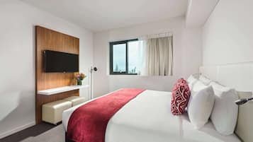 Quarto de hotel moderno com uma cama, uma TV fixada em um painel de madeira, uma janela com cortinas e um pequeno espaço de estar com uma manta vermelha.