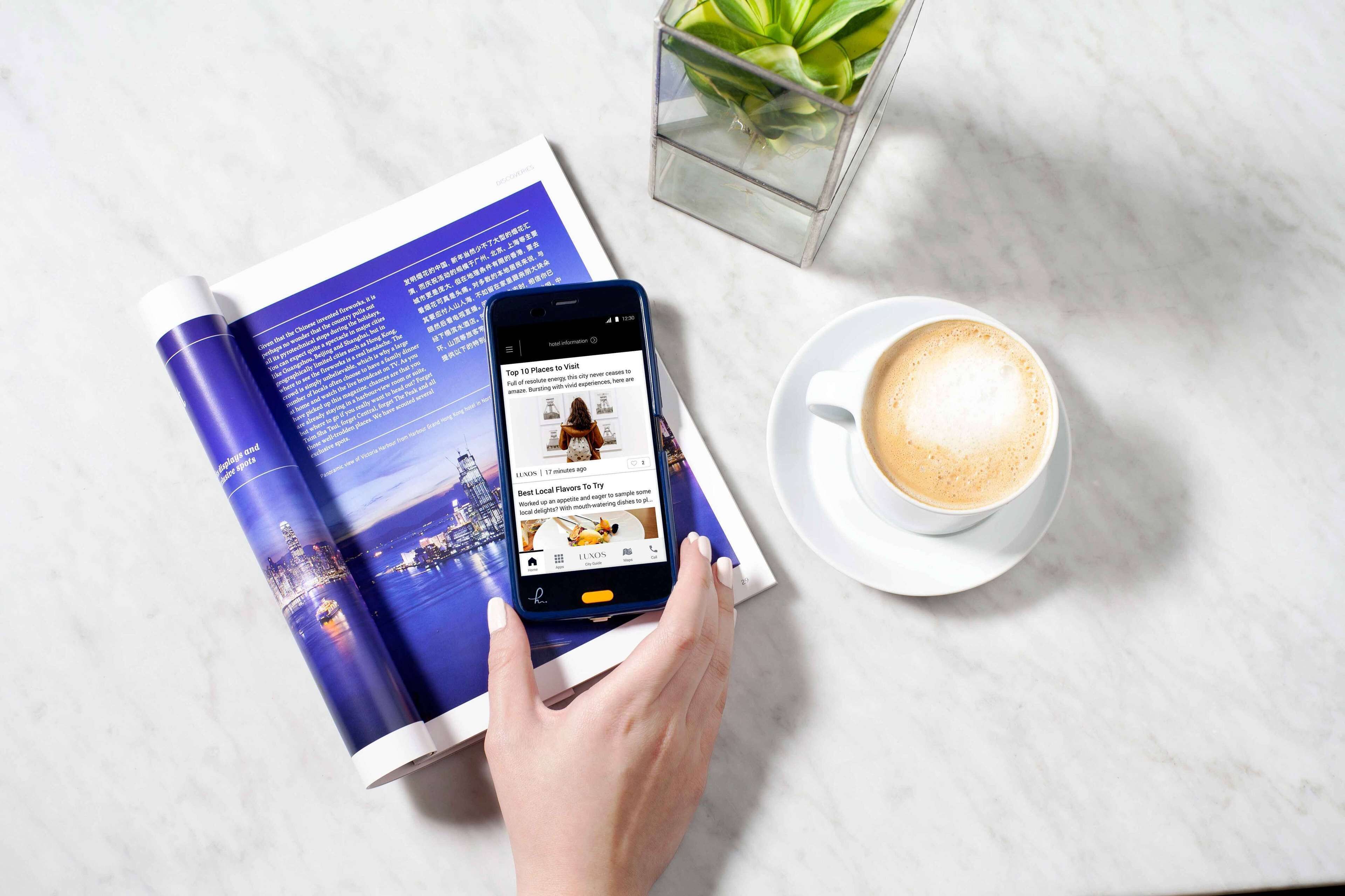 Uma pessoa segurando um smartphone com um aplicativo de viagens aberto, ao lado de um livro e uma xícara de café em uma superfície de mármore.