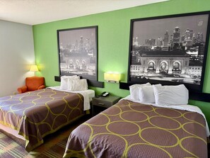Quarto de hotel com duas camas, uma mesa de cabeceira, um abajur e duas fotografias emolduradas em preto e branco de paisagens urbanas na parede.