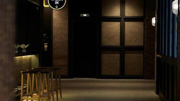 Um corredor com pouca luz, com cadeiras de madeira e um bar à esquerda, uma porta grande no centro e uma parede de tijolos à direita.