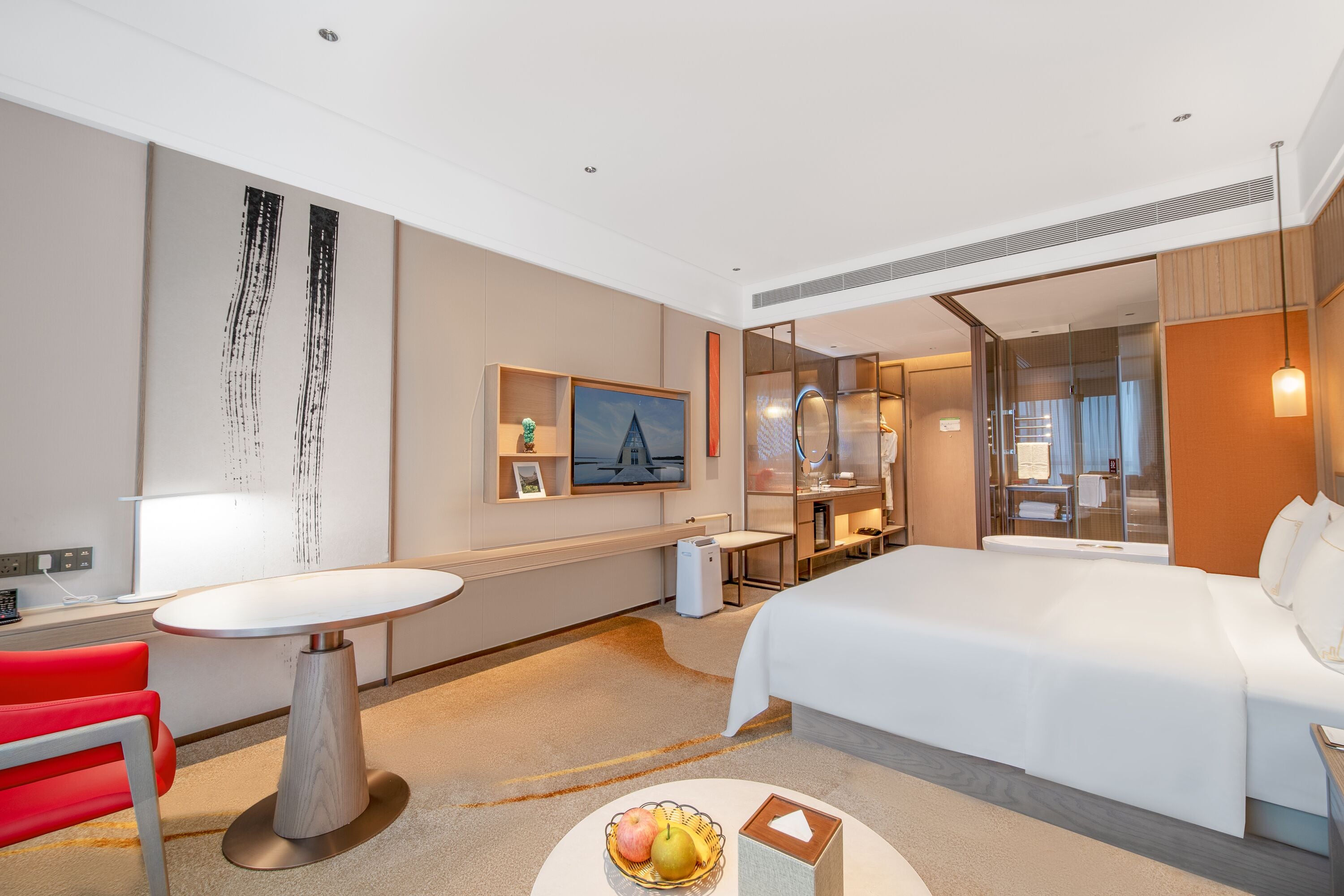 Quarto de hotel moderno com uma cama grande, uma escrivaninha com cadeira, uma mesinha de centro com uma cesta de frutas e uma televisão.