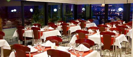 Um restaurante com mesas postas para refeições, amplas janelas e vista para a cidade à noite.