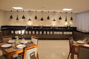 Uma área de refeições moderna em hotel, com um balcão longo, várias mesas e luminárias pendentes.