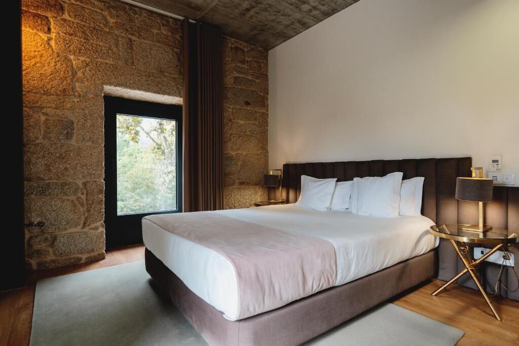 Quarto com parede de pedra, uma cama grande, mesa de cabeceira com abajur e janela com cortinas.