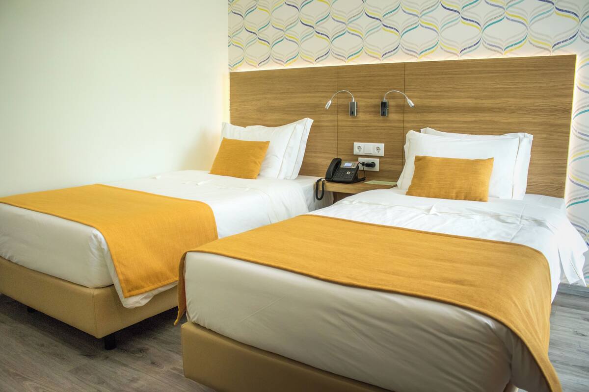 Quarto de hotel com duas camas, cada uma com colcha branca e amarela, cabeceira de madeira e telefone fixado na parede.
