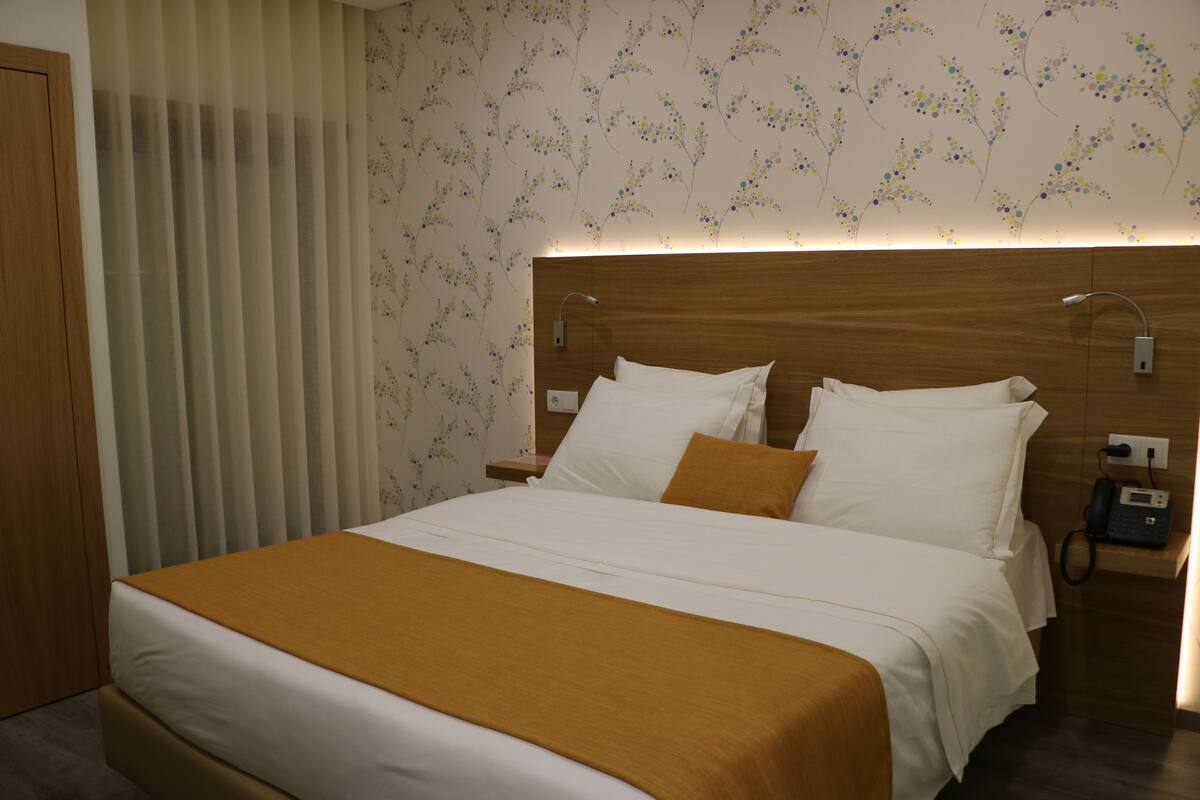 Quarto de hotel com uma cama grande, cabeceira de madeira, mesa de cabeceira com telefone e janela com persianas verticais.
