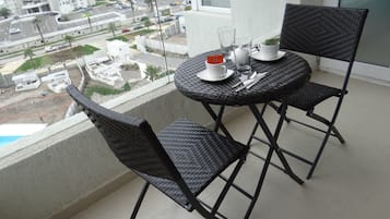 Varanda com duas cadeiras de vime e uma mesa, com vista para um parque de estacionamento e edifícios.