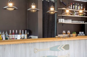 Uma área de bar com prateleiras de madeira que exibem garrafas e copos, iluminação pendente e um enfeite de prancha de remo no balcão.