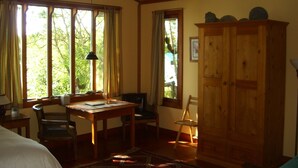 Um quarto com cama, um armário de madeira, uma escrivaninha com cadeira, uma mesa com cadeira e janelas amplas com cortinas.