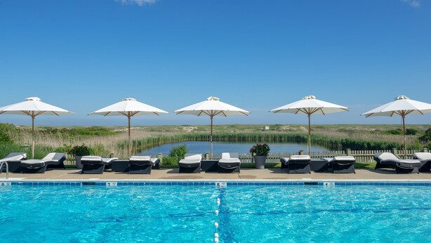 Área da piscina com espreguiçadeiras e guarda-sóis, céu claro e vista para a praia e dunas ao fundo.
