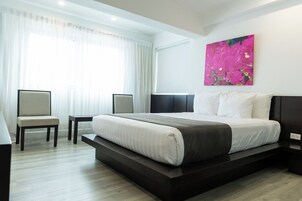 Quarto de hotel moderno com uma cama grande, duas cadeiras, uma mesa de cabeceira e uma janela ampla com cortinas finas.