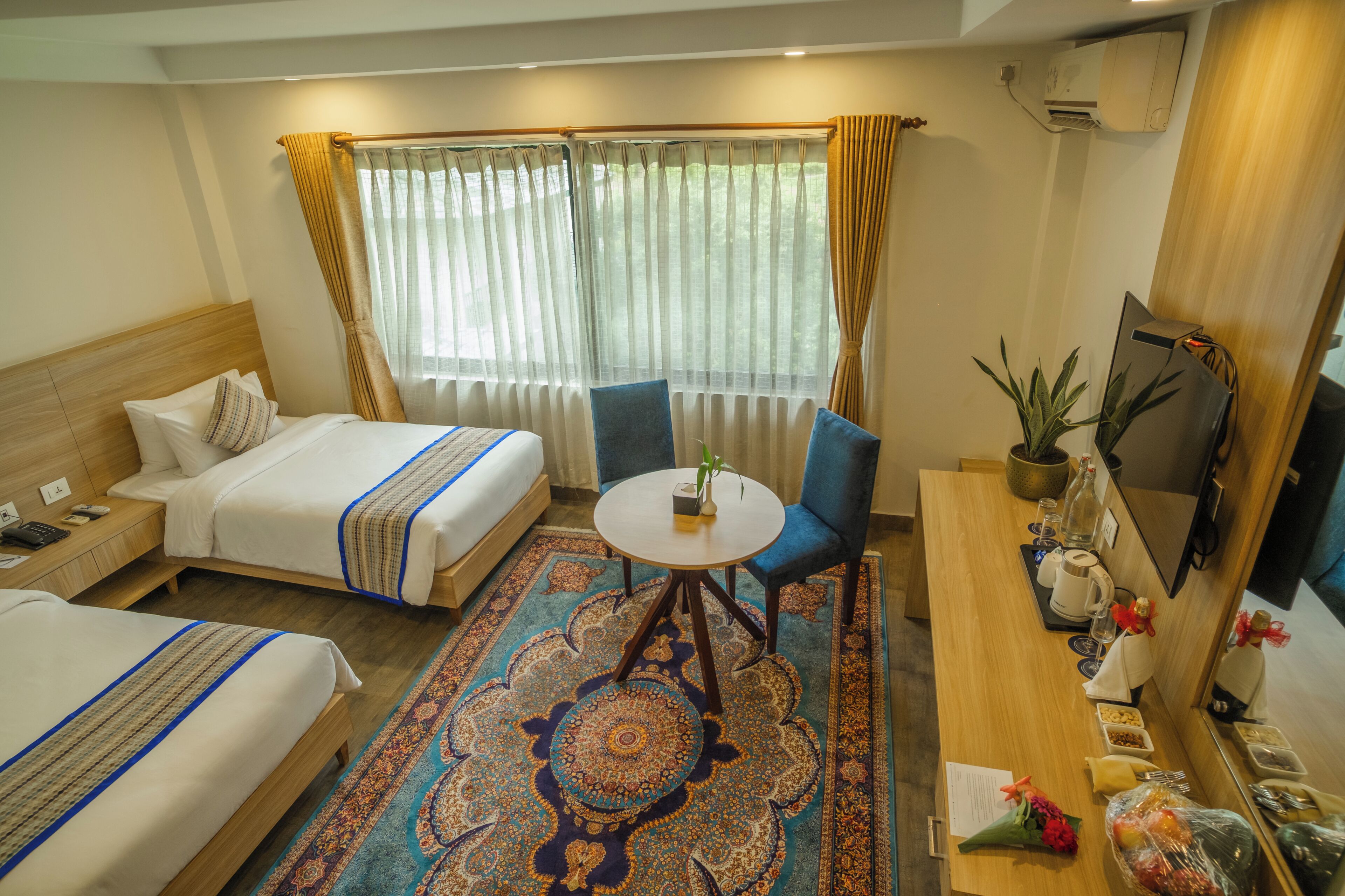 Quarto de hotel com duas camas, uma mesa de jantar com cadeiras, televisão e janela com cortinas.