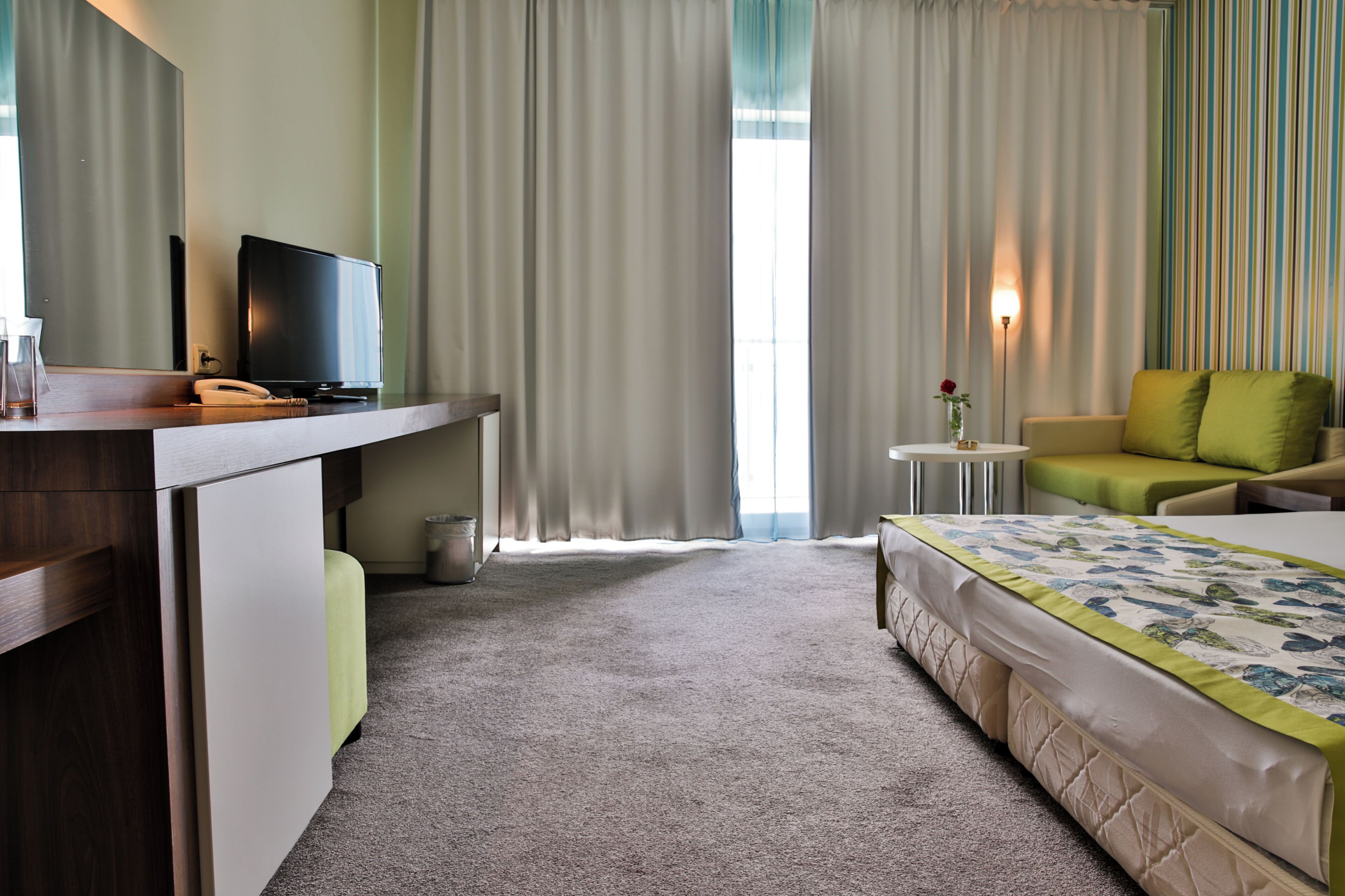 Quarto de hotel com uma cama, uma escrivaninha com televisão, uma mesinha com um vaso e uma janela com cortinas.