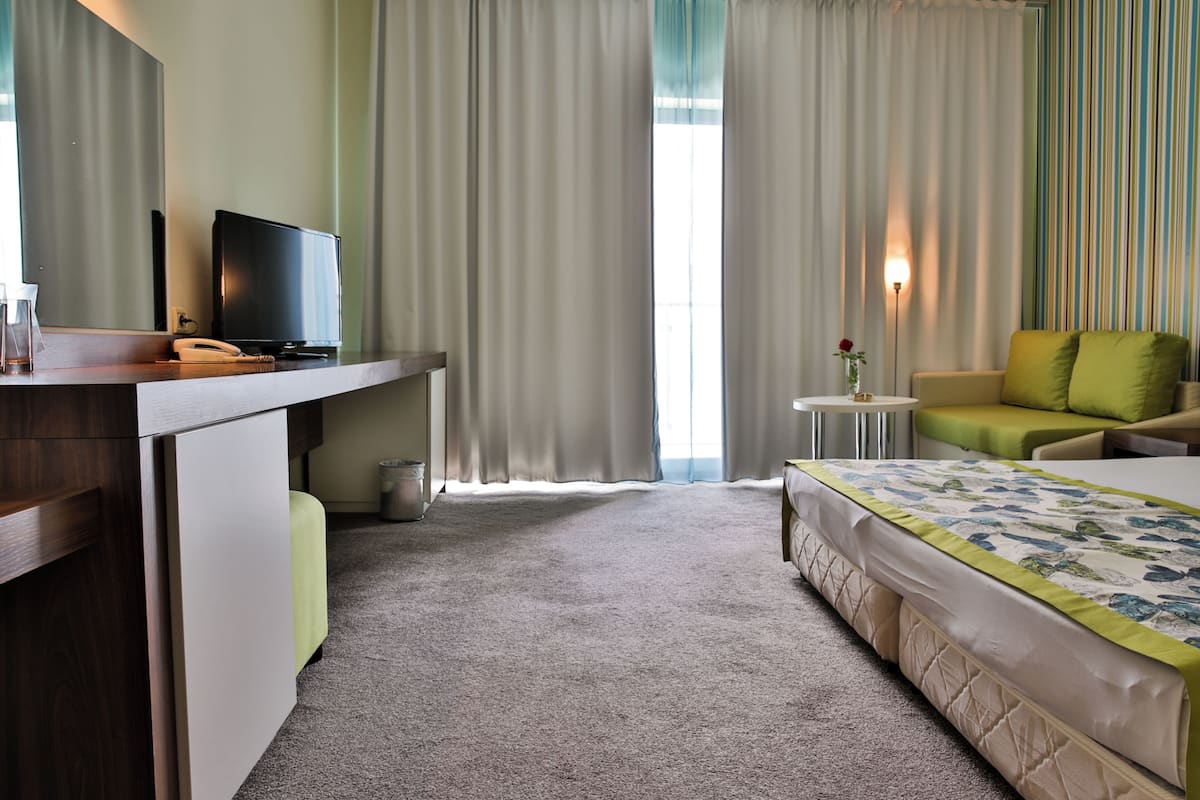 Quarto de hotel com uma cama, uma escrivaninha com televisão, uma mesinha com um vaso e uma janela com cortinas.