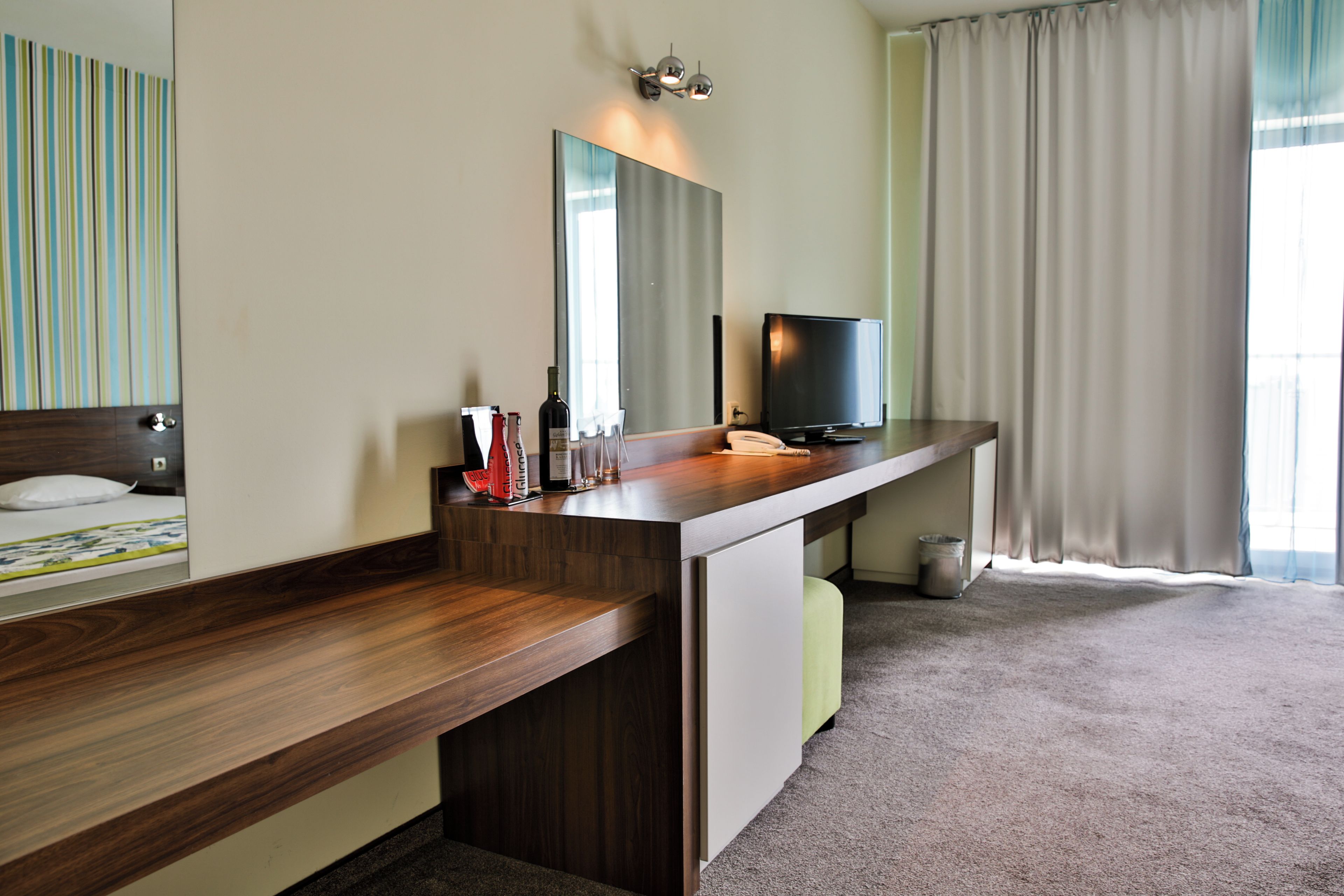 Quarto de hotel com escrivaninha de madeira, televisão de tela plana, espelho grande, cama com travesseiro e janela com cortinas.