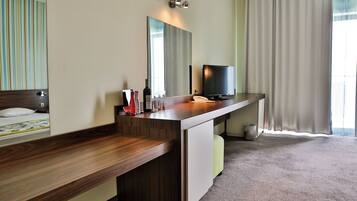 Quarto de hotel com escrivaninha de madeira, televisão de tela plana, espelho grande, cama com travesseiro e janela com cortinas.