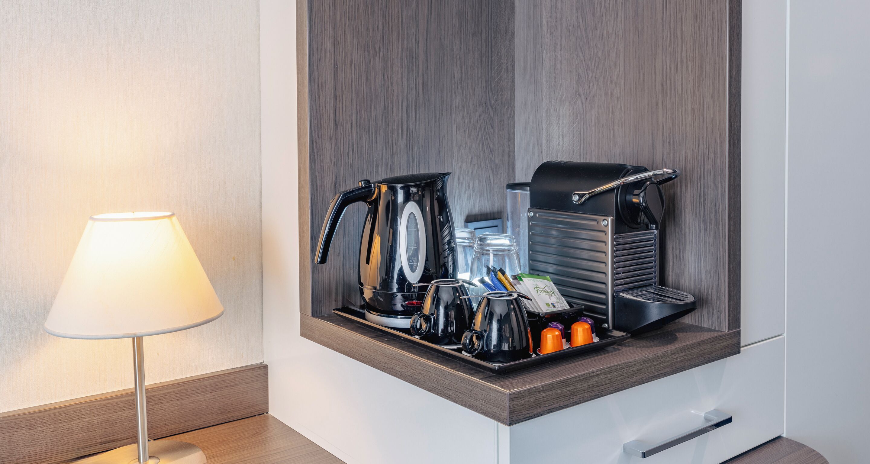 Uma kitchenette compacta com cafeteira, chaleira e chávenas laranja numa bandeja.