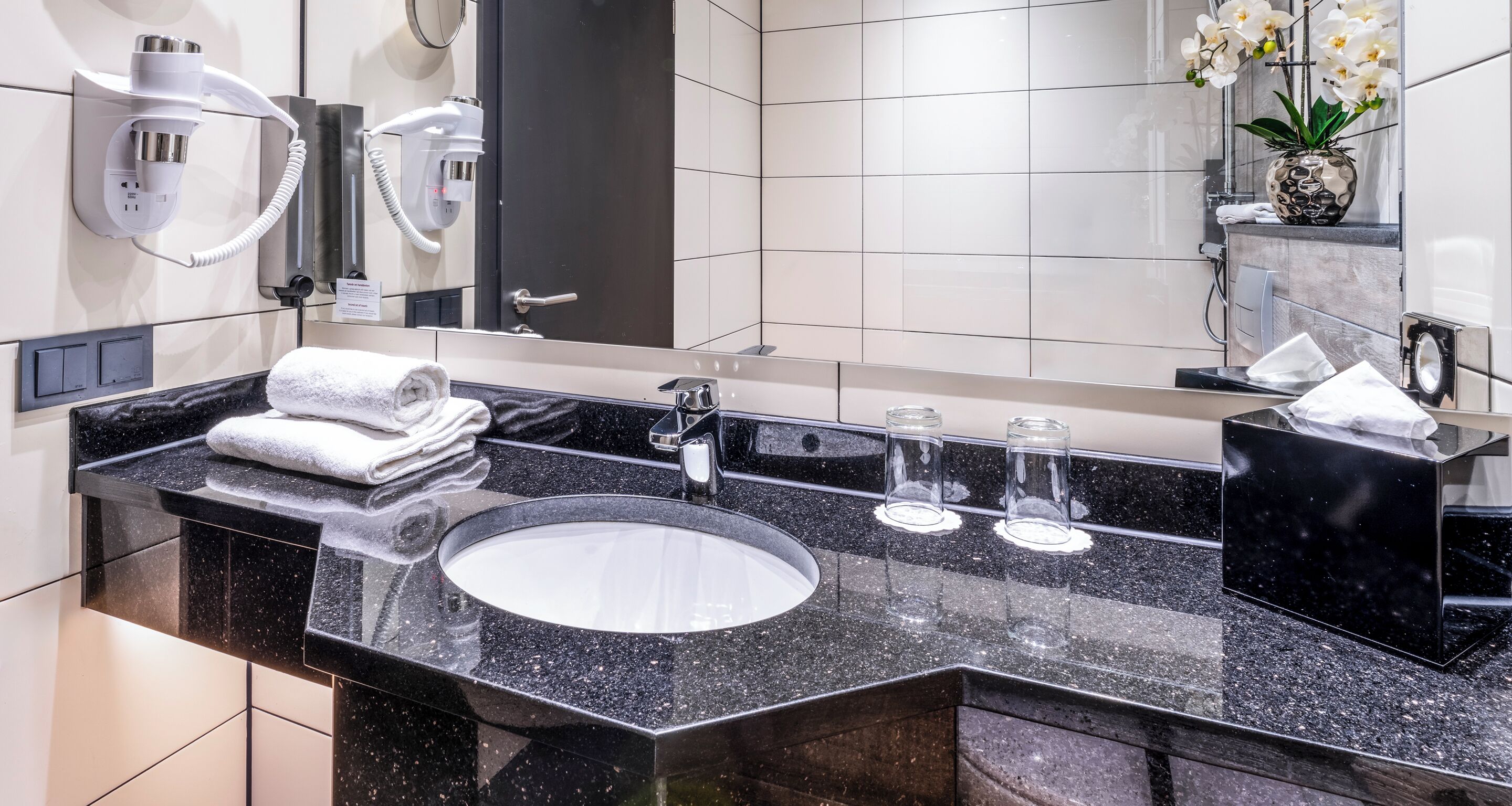 Casa de banho moderna com bancada de granito preto, lavatório redondo e dois secadores de cabelo fixados na parede.