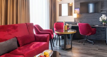 Quarto de hotel moderno com sofá vermelho, poltronas a combinar, mesa de jantar redonda com champanhe e copos, e televisão na parede.