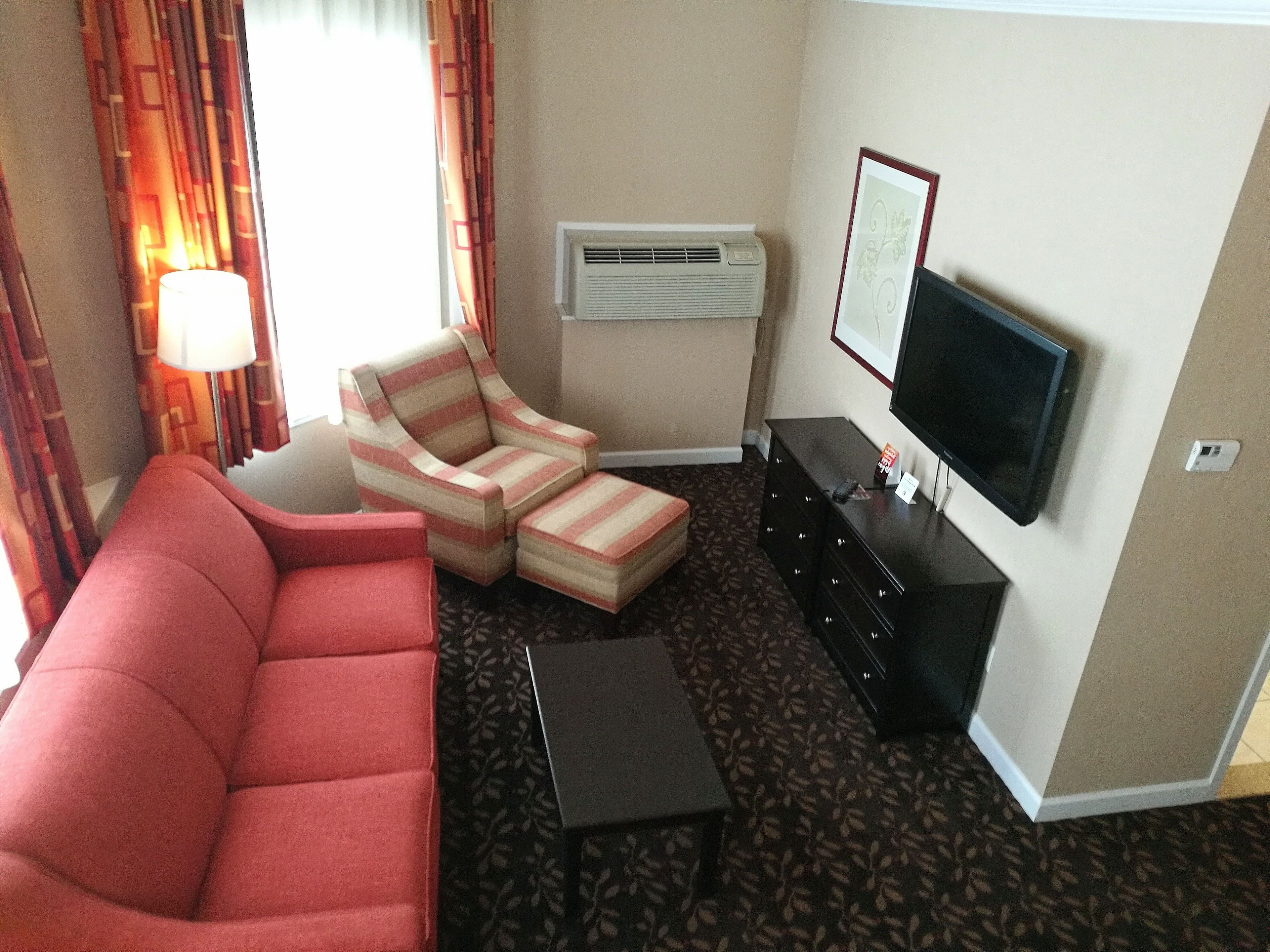 Quarto de hotel com sofá vermelho, poltrona listrada, pequena mesa, TV e aparelho de ar condicionado.
