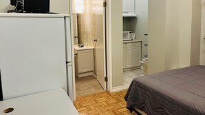 Quarto compacto com cama, escrivaninha, computador, pequeno espaço para refeições e banheiro.