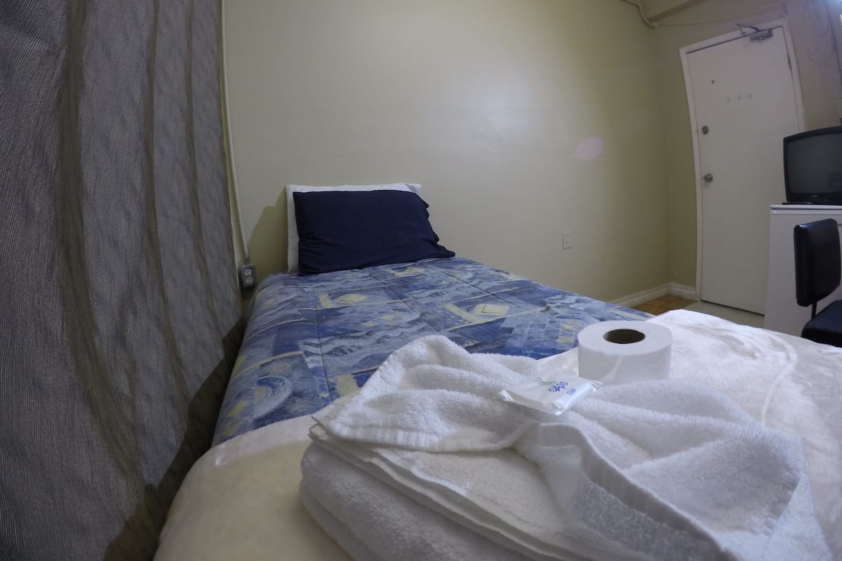 Um quarto pequeno e simples, com uma cama, uma escrivaninha, uma cadeira e uma televisão.
