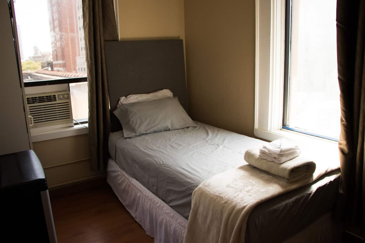Uma cama de solteiro com cabeceira cinza, uma janela com cortinas e uma pilha de toalhas dobradas no rodapé da cama.