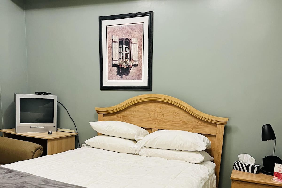 Cama bem arrumada com lençóis brancos, cabeceira de madeira, quadro na parede, mesa de cabeceira com abajur e uma pequena TV em suporte de madeira.