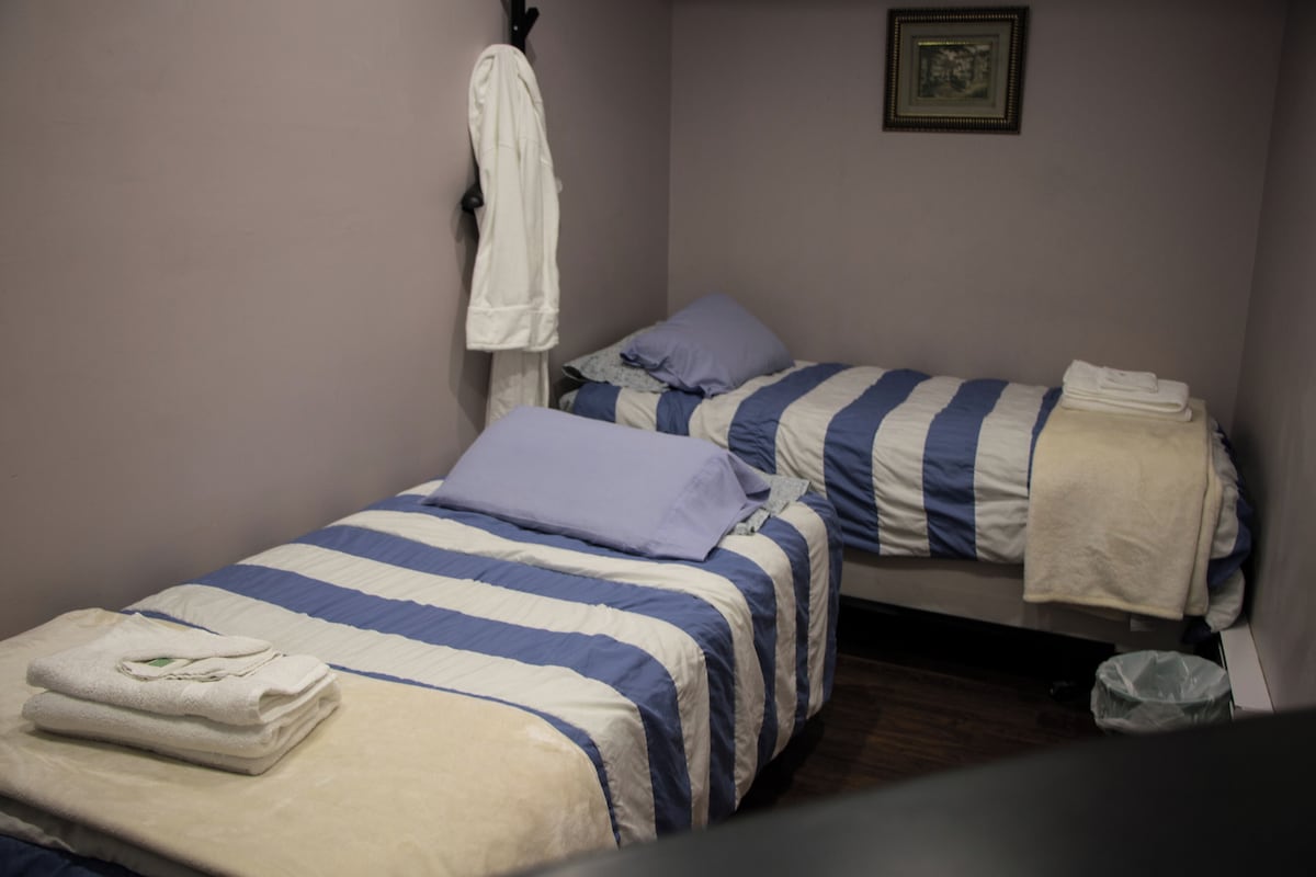 Quarto de hotel pequeno e individual com duas camas, cada uma com roupa de cama listrada. Há um roupão branco pendurado em um gancho e um quadro na parede.
