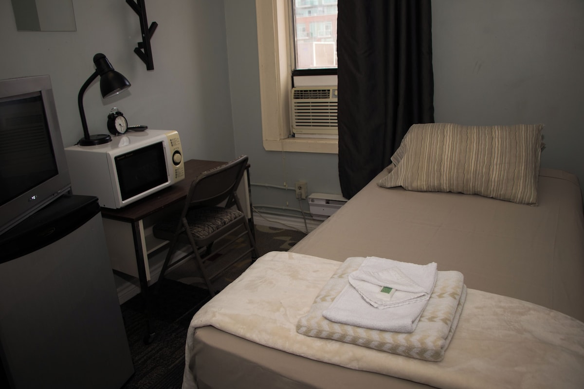 Quarto de hotel pequeno com cama, escrivaninha com computador, micro-ondas e janela com cortinas.