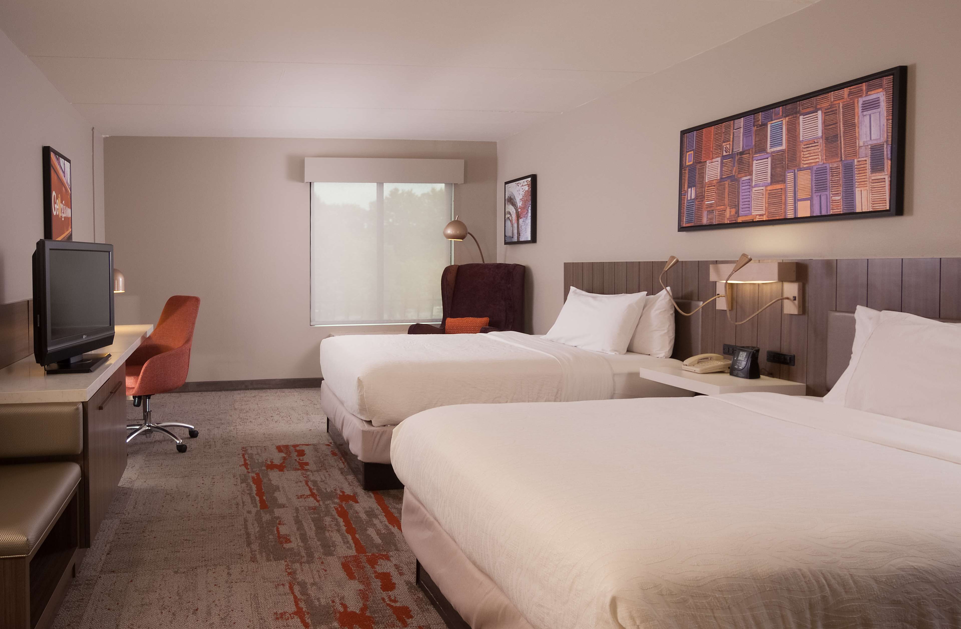 Quarto de hotel com duas camas, uma escrivaninha com computador, uma cadeira, televisão, janela com persianas e um quadro na parede.