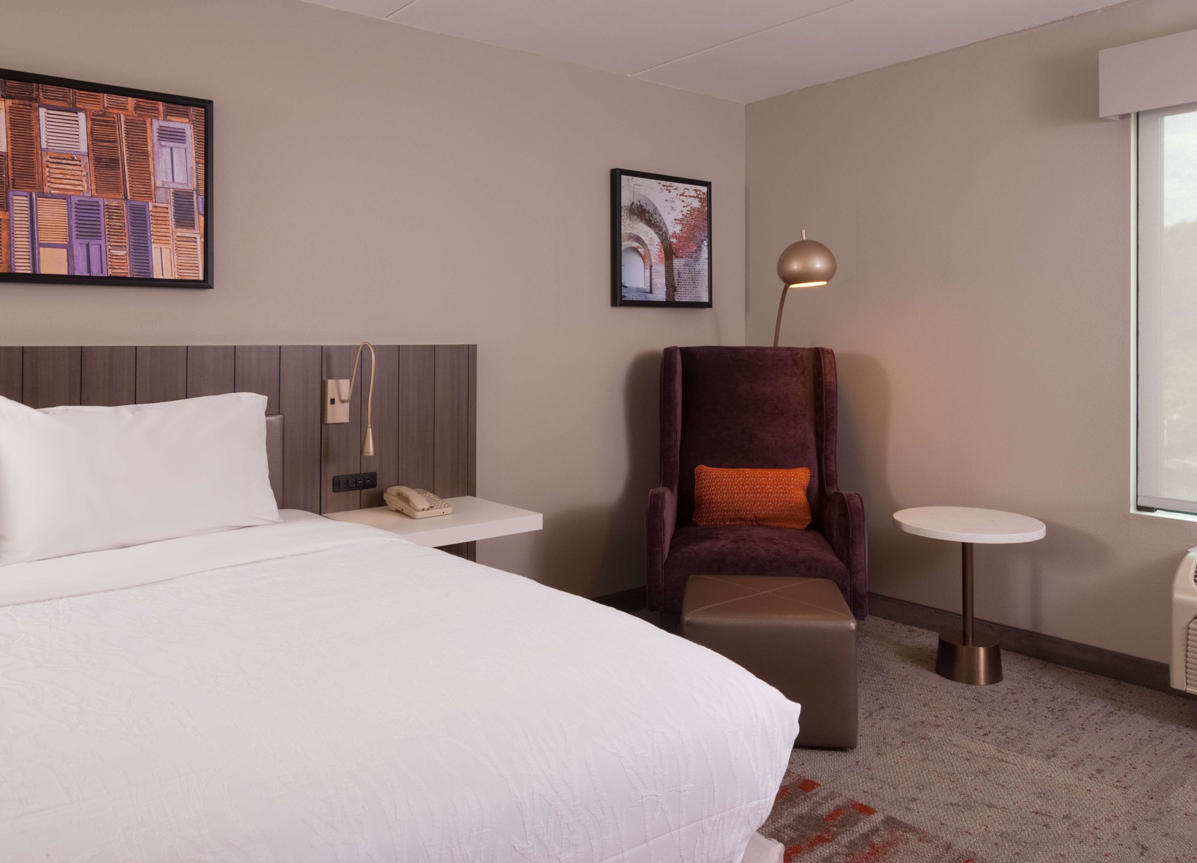Quarto de hotel com uma cama grande, uma cadeira, uma mesinha, um abajur, um telefone e obras de arte nas paredes.