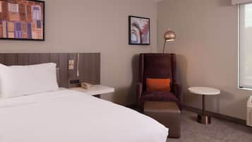 Quarto de hotel com uma cama grande, uma cadeira, uma mesinha, um abajur, um telefone e obras de arte nas paredes.
