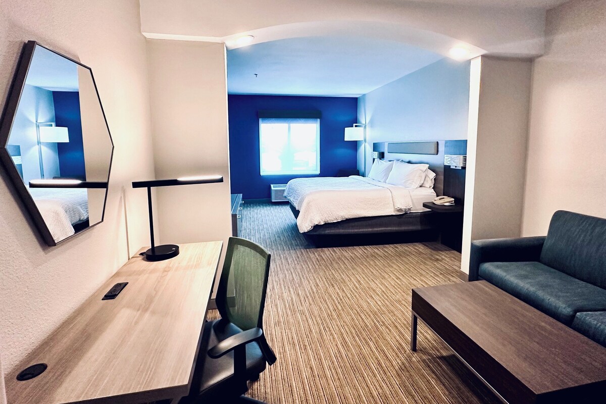 Quarto de hotel com cama, escrivaninha, cadeira, sofá, espelho e janela.