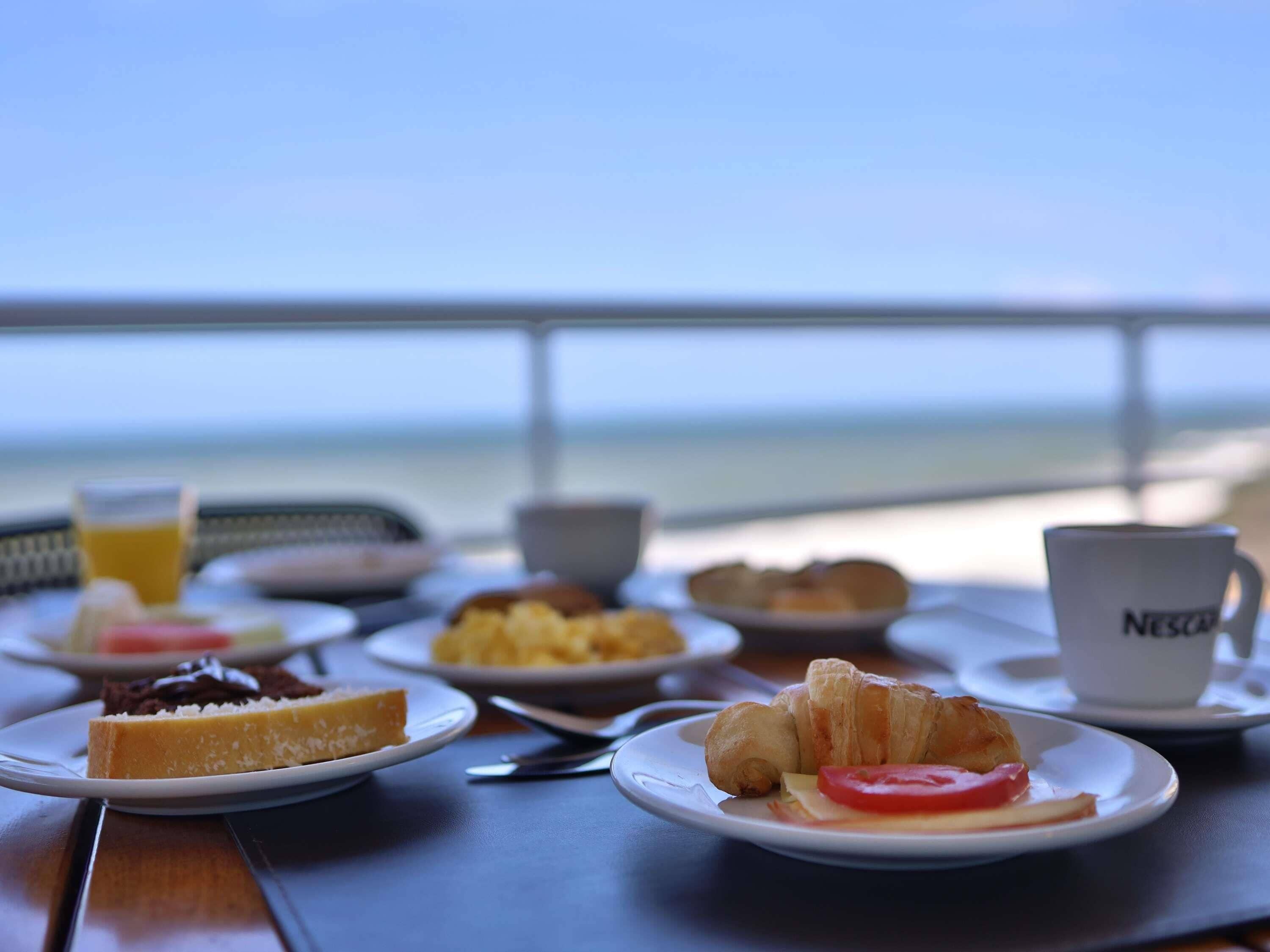 Café da manhã com croissants, uma fatia de tomate e uma xícara de café sobre uma mesa com vista para a praia.