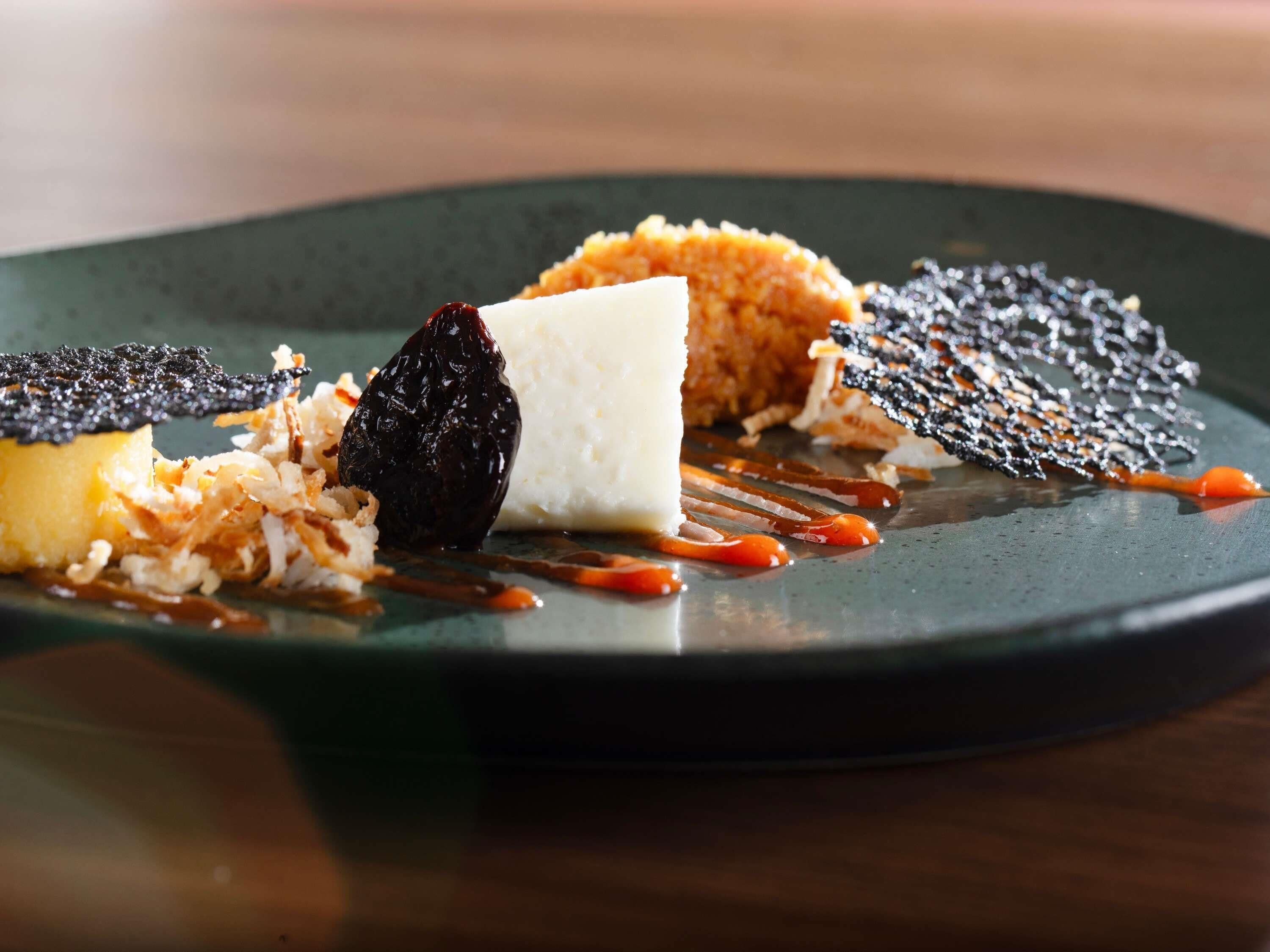 Um prato de sobremesa com diversos elementos, incluindo uma fruta escura, um creme branco e um fio de caramelo.