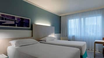 Quarto de hotel com duas camas, uma escrivaninha, uma cadeira, um quadro na parede e uma janela com cortinas.