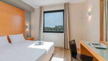 Quarto de hotel com uma cama grande, uma escrivaninha, uma cadeira, uma janela com cortinas e vista para árvores.