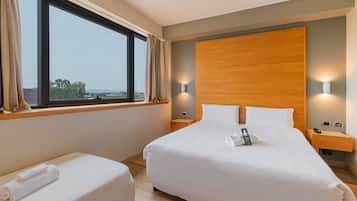 Quarto de hotel com uma cama grande, uma cama menor, uma janela com cortinas, cabeceira de madeira, mesas de cabeceira com luminárias e vista para a cidade.