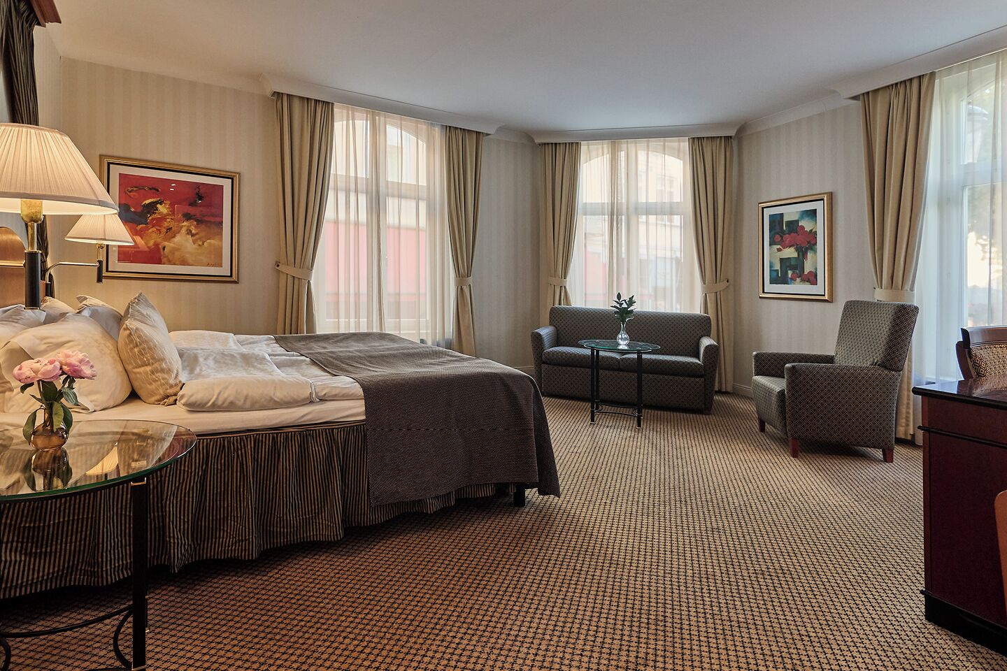 Quarto de hotel com cama, sofá, poltrona, mesa pequena e luminária.